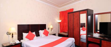 Quarto de hotel com uma cama grande, um guarda-roupa, uma escrivaninha com espelho, uma televisão e um telefone numa mesinha de cabeceira.