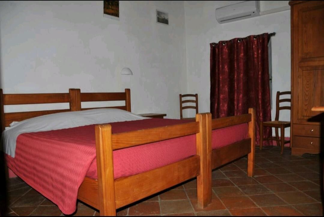 Quarto individual com uma cama de madeira, colcha vermelha, mesa de cabeceira pequena, uma cadeira e um guarda-roupa.