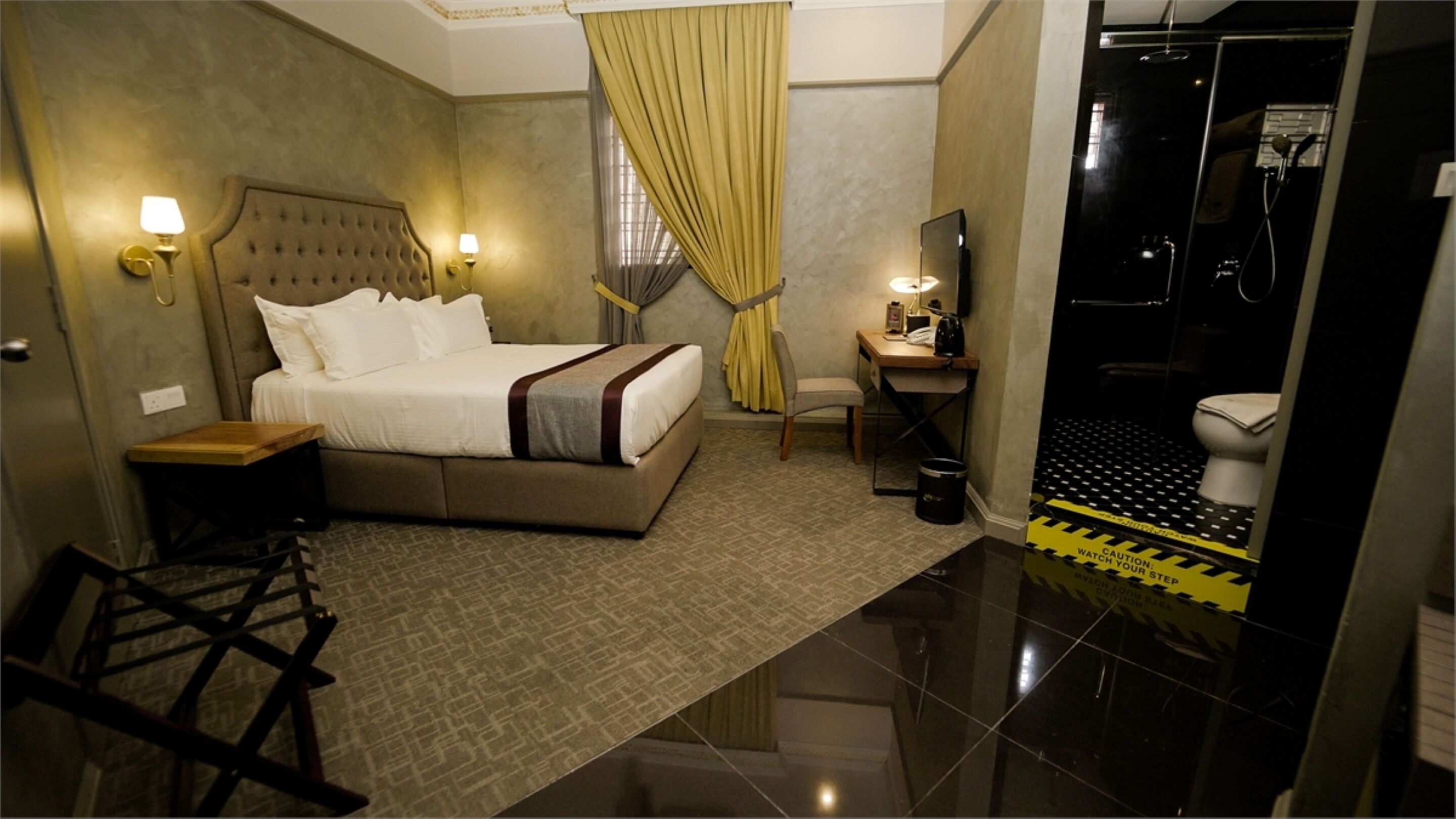 Quarto de hotel com cama, abajures, escrivaninha, cadeira e banheiro com chuveiro e vaso sanitário.