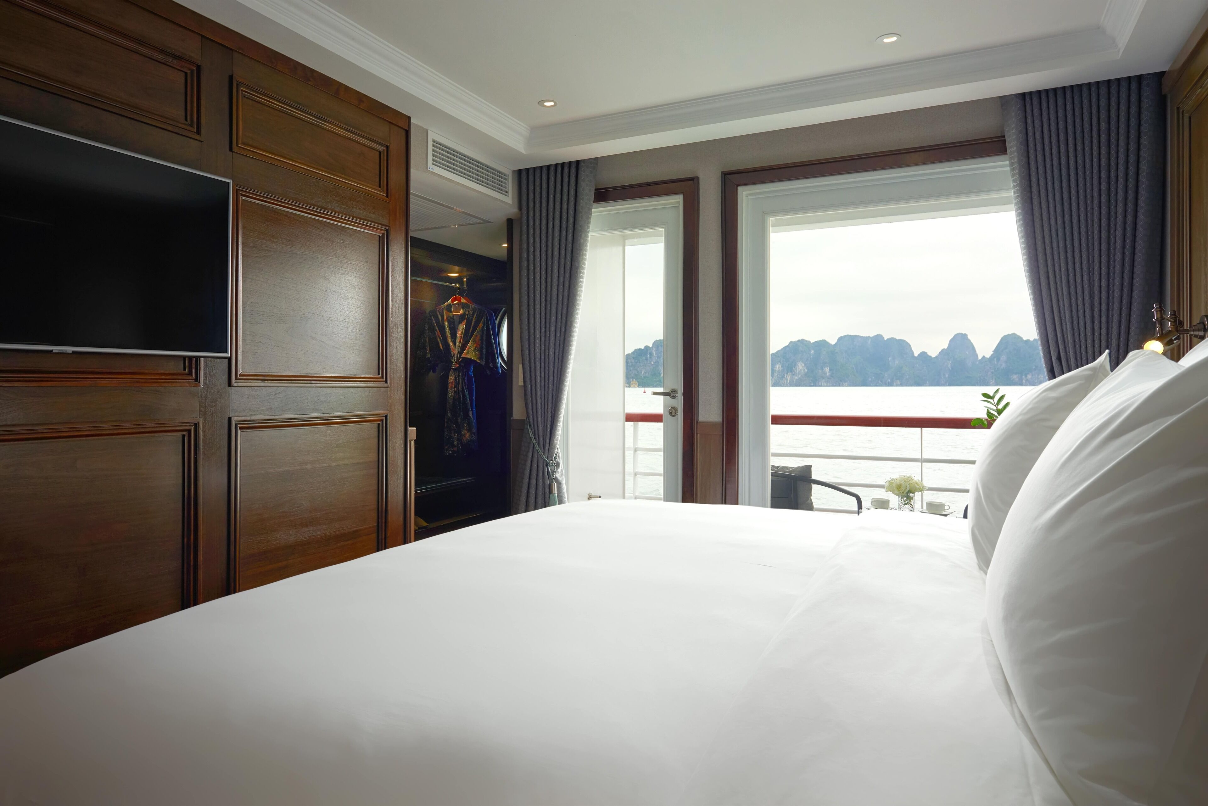 Quarto de hotel com uma cama grande, uma televisão fixada na parede e vista para o mar através de uma janela ampla.