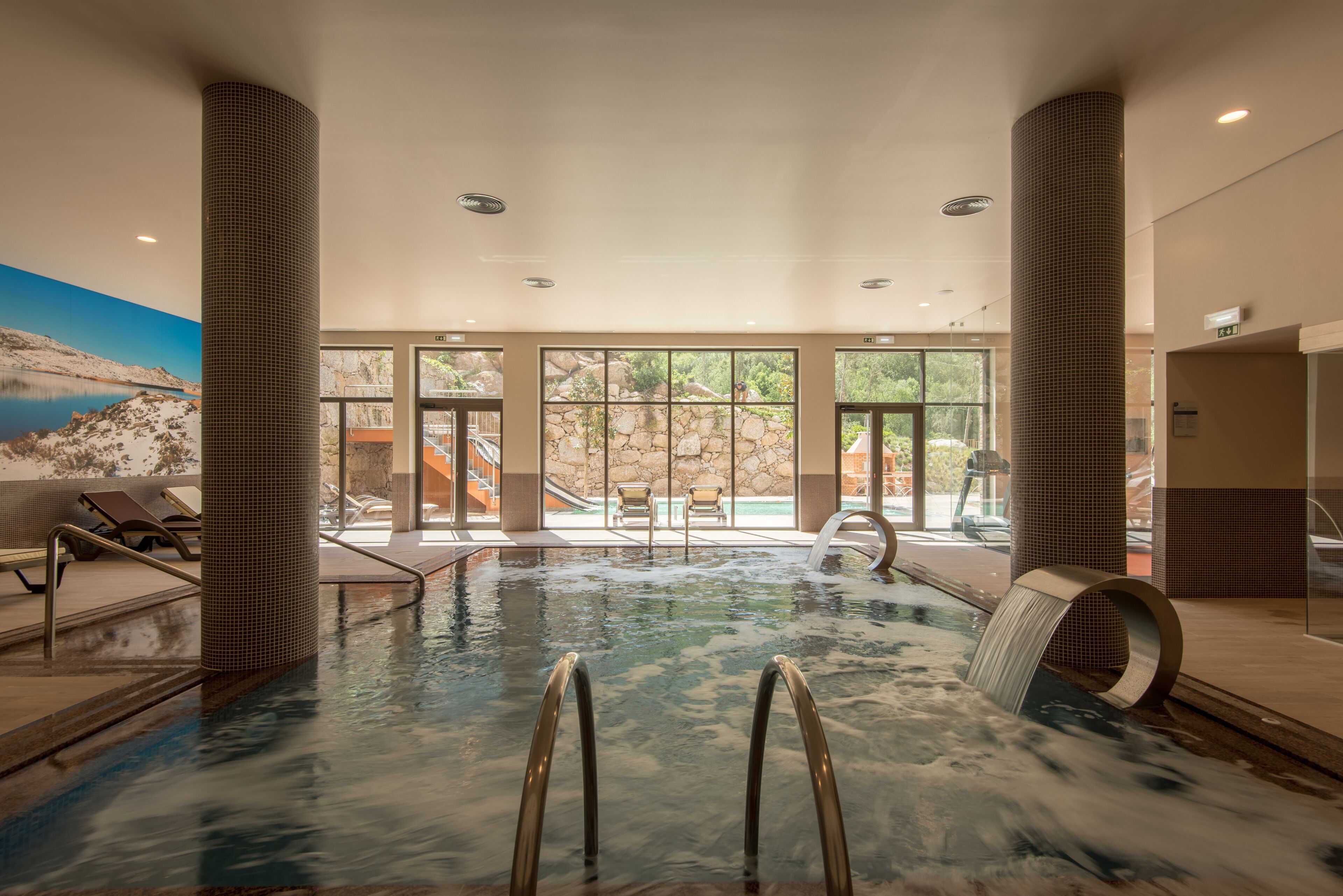 Piscina interior com água cristalina, vista para o exterior e parede de pedra.