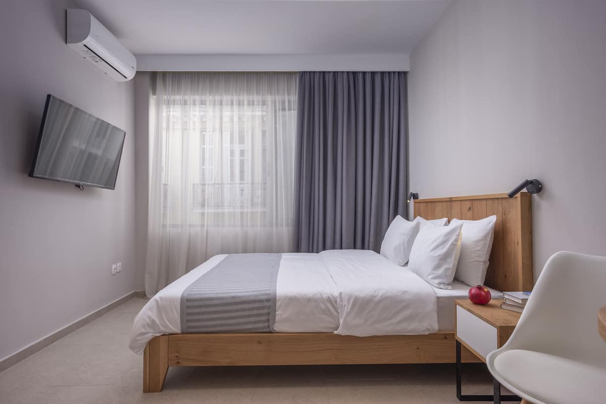 Quarto de hotel moderno com uma cama de madeira, televisão de tela plana na parede, mesa de cabeceira com abajur e uma janela grande com cortinas finas.