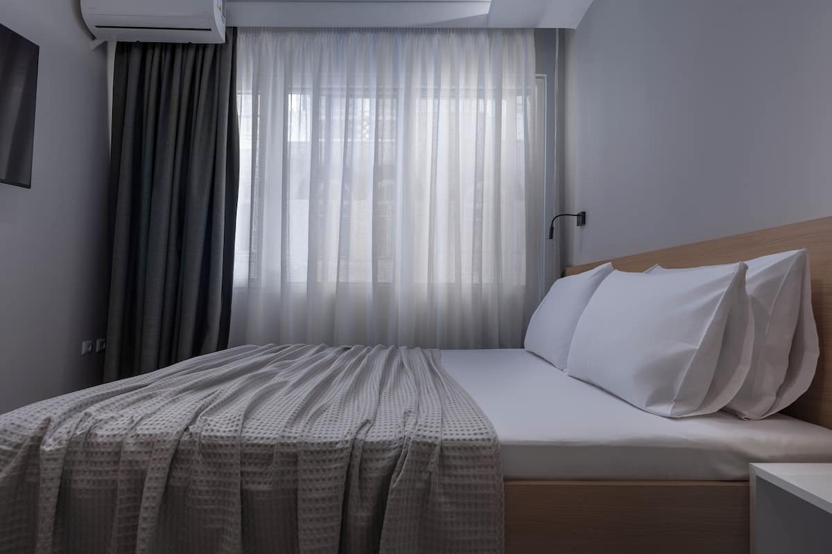 Uma cama bem arrumada com lençóis brancos, cabeceira de madeira e janela com cortinas finas.