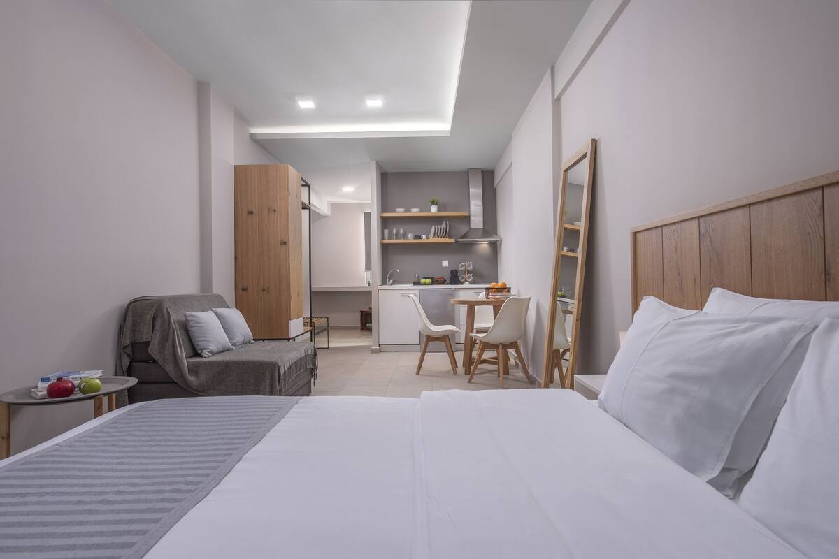 Quarto de hotel pequeno e funcional, com cama, mesinha, cadeira, cozinha compacta e armário.