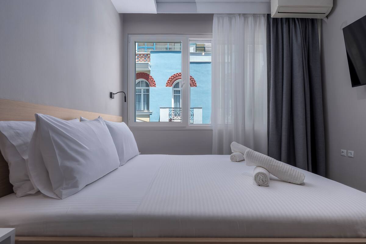 Uma cama bem arrumada com lençóis brancos, uma janela com vista para um prédio e um ar-condicionado fixado na parede.