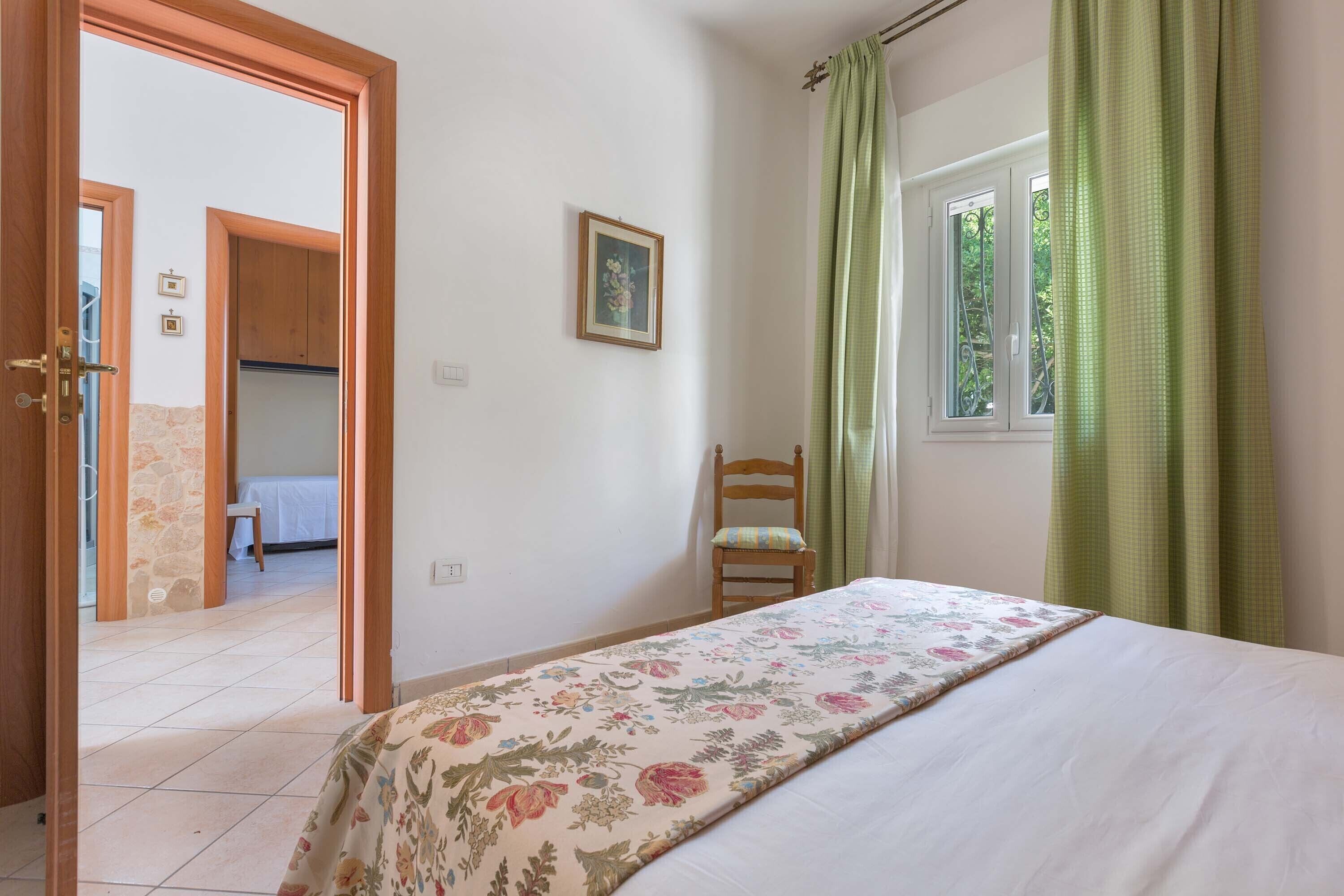 Quarto com cama, cadeira, quadro na parede e janela com cortinas.