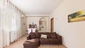 Uma sala de estar com um sofá seccional marrom, uma parede branca com um quadro emoldurado e uma área de cozinha ao fundo.