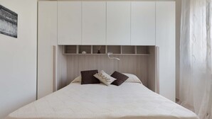 Quarto com cama, cabeceira com prateleiras e luminária fixada na parede.