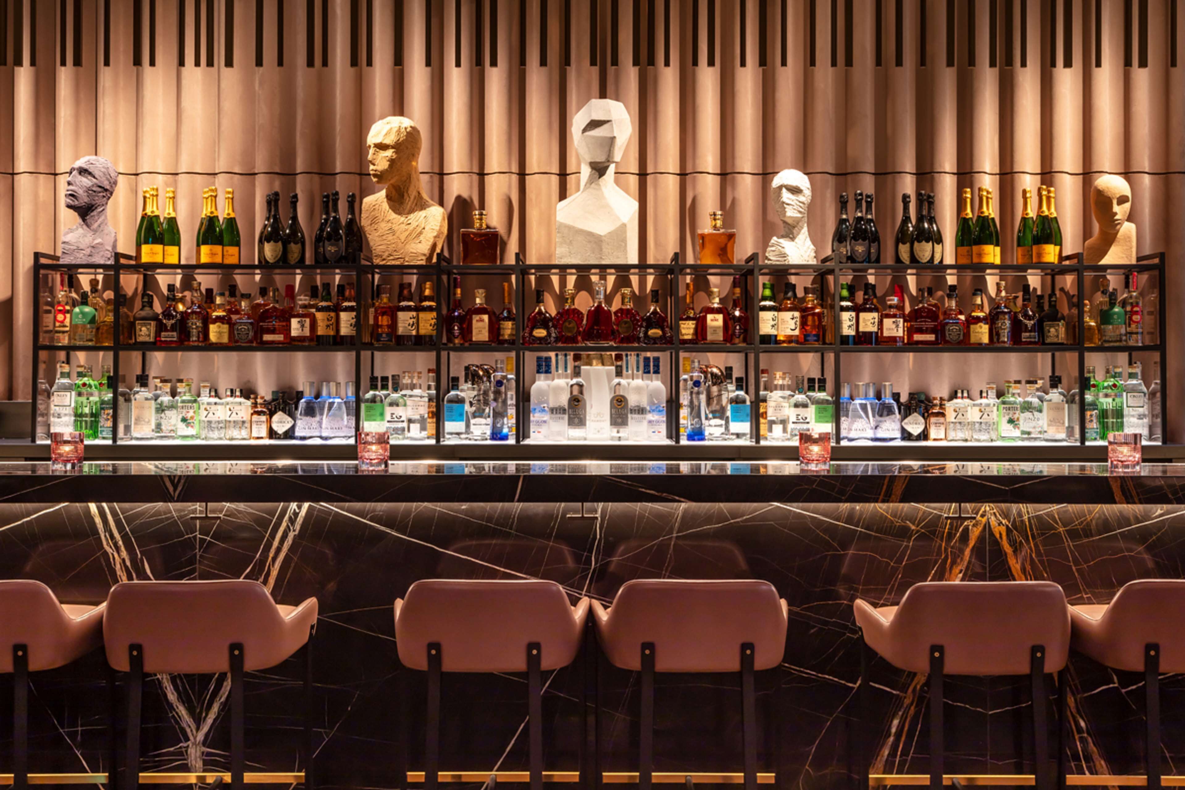 Um bar com várias prateleiras de garrafas de bebidas, uma escultura central e banquetas.