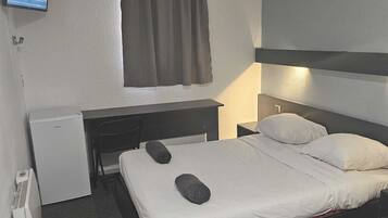 Quarto de hotel com cama, mesa de trabalho, cadeira, televisão fixada na parede e janela com cortinas.