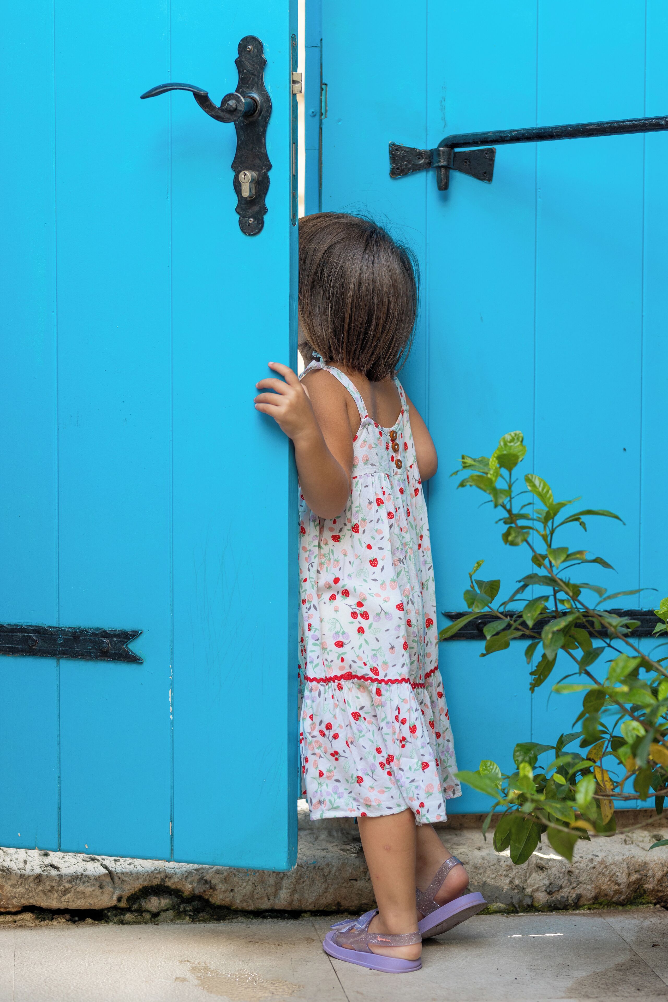 Criança com vestido floral parada em frente a uma porta azul com puxador de metal.