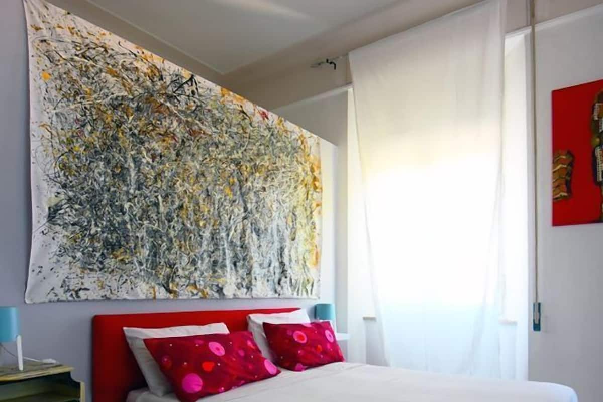 Quarto com cama vermelha, roupa de cama branca e uma grande pintura abstrata na parede.