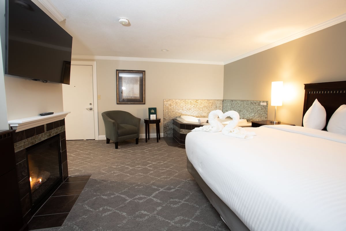 Quarto de hotel com cama, televisão na parede, lareira, poltrona, mesa pequena e banheiro com pia e espelho.