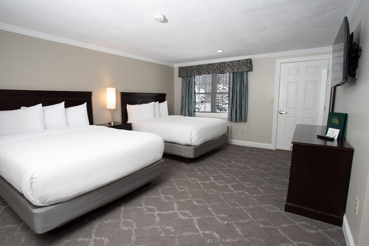 Quarto de hotel com duas camas, uma escrivaninha, uma televisão e uma janela com cortinas.