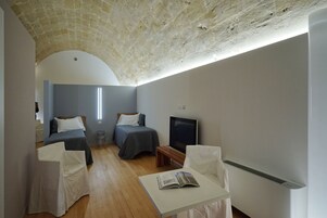 Quarto com teto alto abobadado, duas camas com cabeceiras, uma televisão, uma mesinha com um livro e um banquinho.