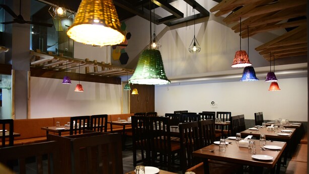 Restaurante com mesas e cadeiras de madeira, luminárias pendentes e parede branca com decoração geométrica.