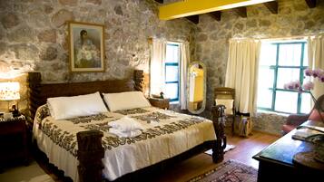 Quarto com paredes de pedra, uma cama grande, cabeceira de madeira, mesa de cabeceira com luminária e um espelho na parede.