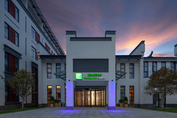 Entrada moderna de um hotel com porta de vidro, placa da Holiday Inn Express e céu azul ao fundo.