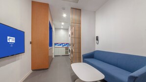 Quarto de hotel com sofá azul, uma pequena mesa branca, porta de madeira e parede azul com um logotipo.