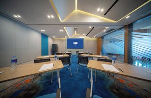 Sala de conferências com mesas, cadeiras, tela de projetor e bebedouro.