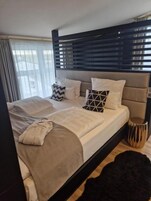 Quarto de hotel moderno com cama, cabeceira, mesa de cabeceira e janela com cortinas.