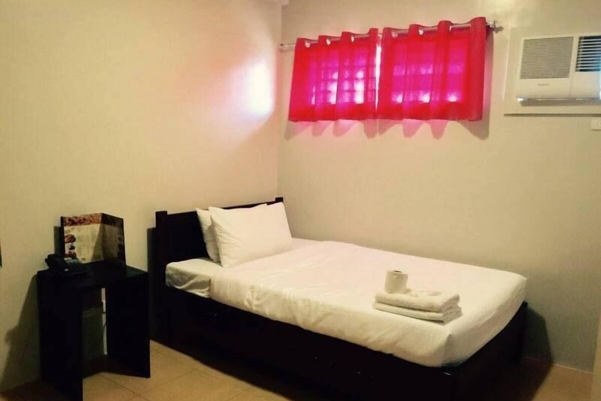 Uma cama de solteiro com roupa de cama branca, uma pequena mesa de cabeceira, uma cortina vermelha e um ar-condicionado fixado na parede.
