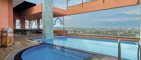 Piscina infinita no terraço com vista clara da paisagem urbana, rodeada por espreguiçadeiras e um deck de madeira.