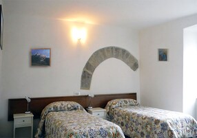 Quarto de hotel com duas camas, uma mesa de cabeceira, uma luminária fixa na parede e um arco decorativo de pedra.
