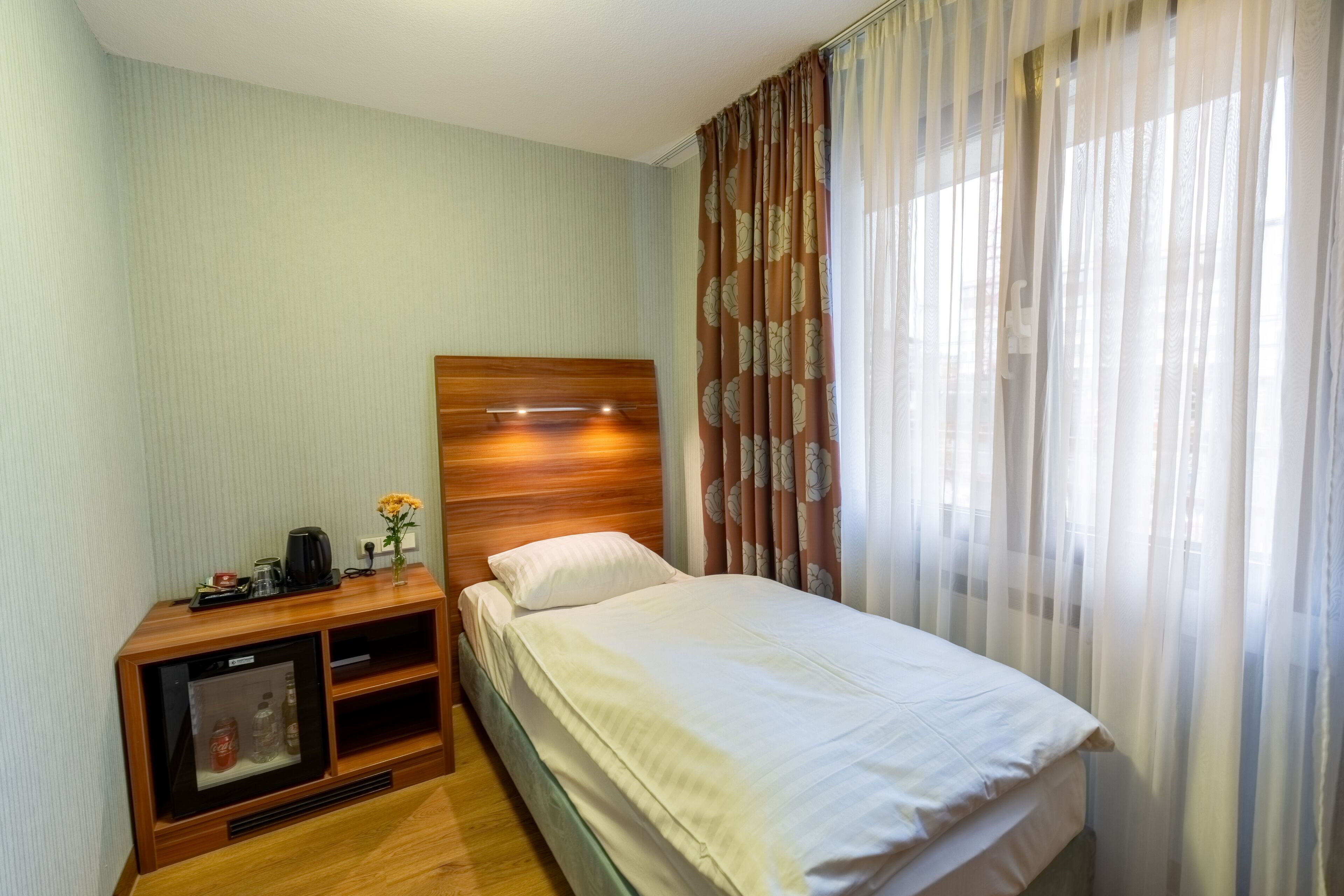 Quarto de hotel com cabeceira de madeira, cama com roupa de cama branca, pequena mesa de cabeceira de madeira com cafeteira e mini-frigorífico.