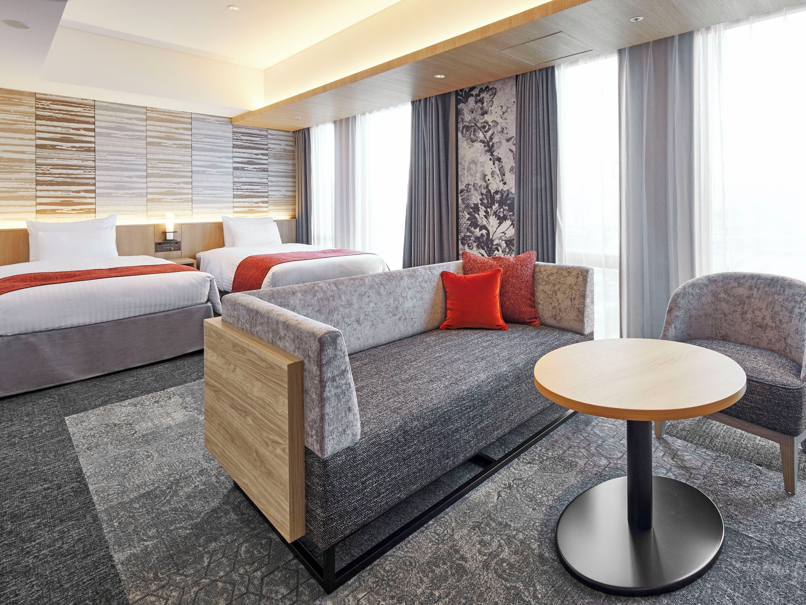 Quarto de hotel moderno com sofá, duas camas, uma mesa redonda e uma janela grande com cortinas.