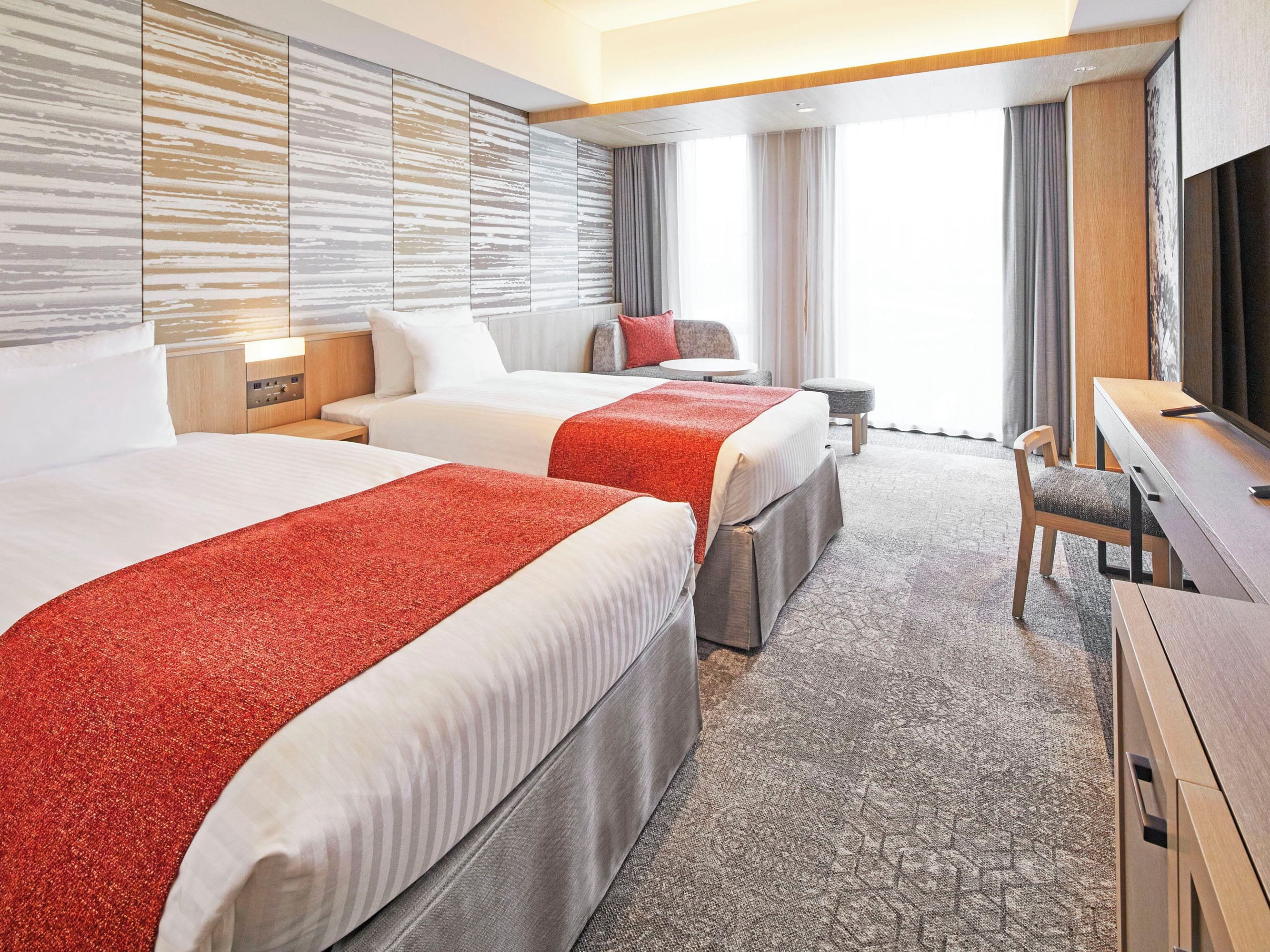 Quarto de hotel com duas camas, uma escrivaninha, uma cadeira e uma janela grande.