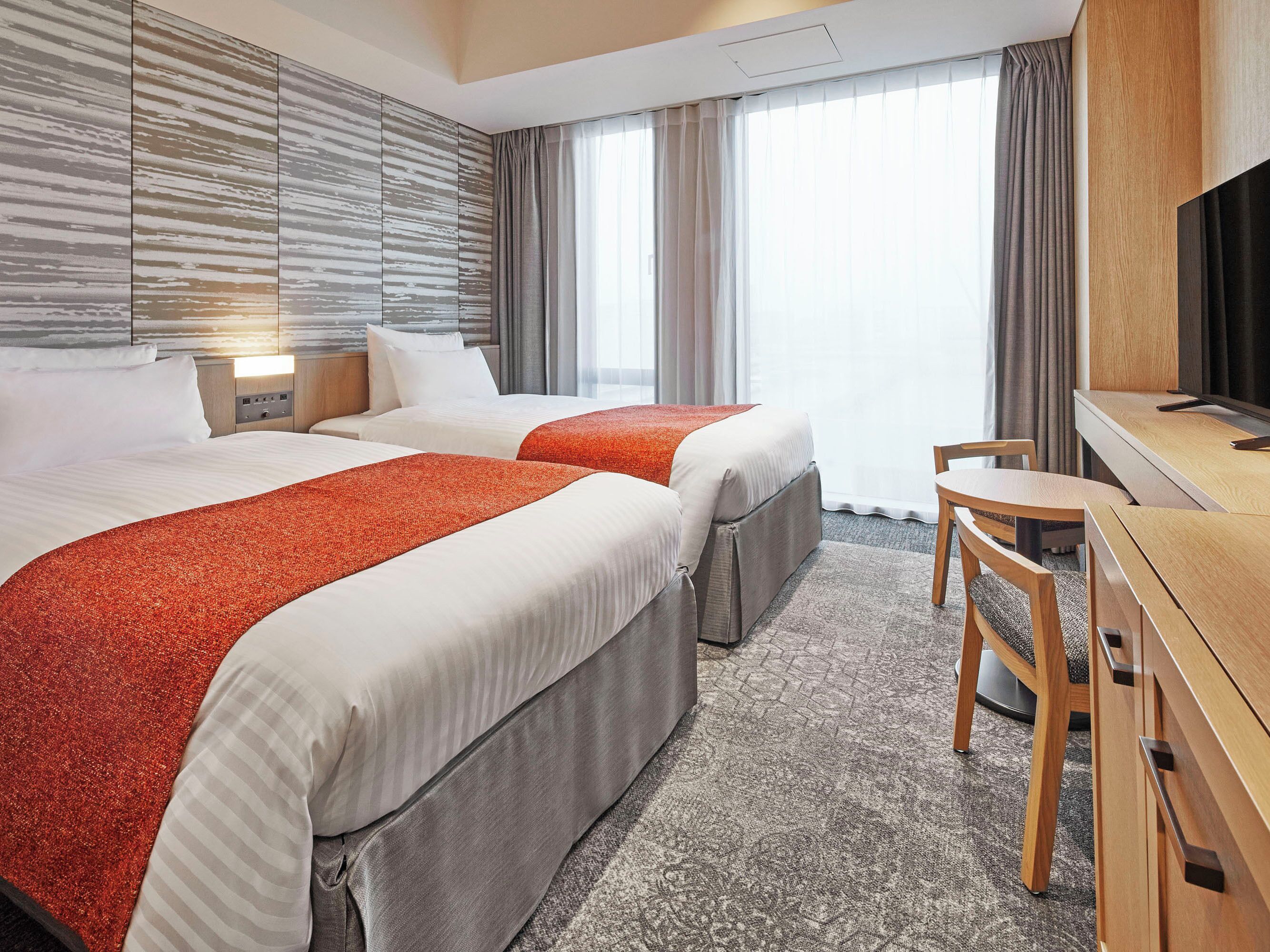 Quarto de hotel com duas camas, uma escrivaninha, uma cadeira, uma televisão e uma janela grande com cortinas.