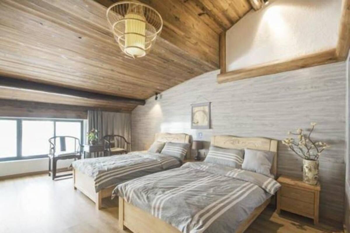 Quarto com duas camas, piso de madeira, luminária no teto, uma janela e mesa de cabeceira com um vaso.