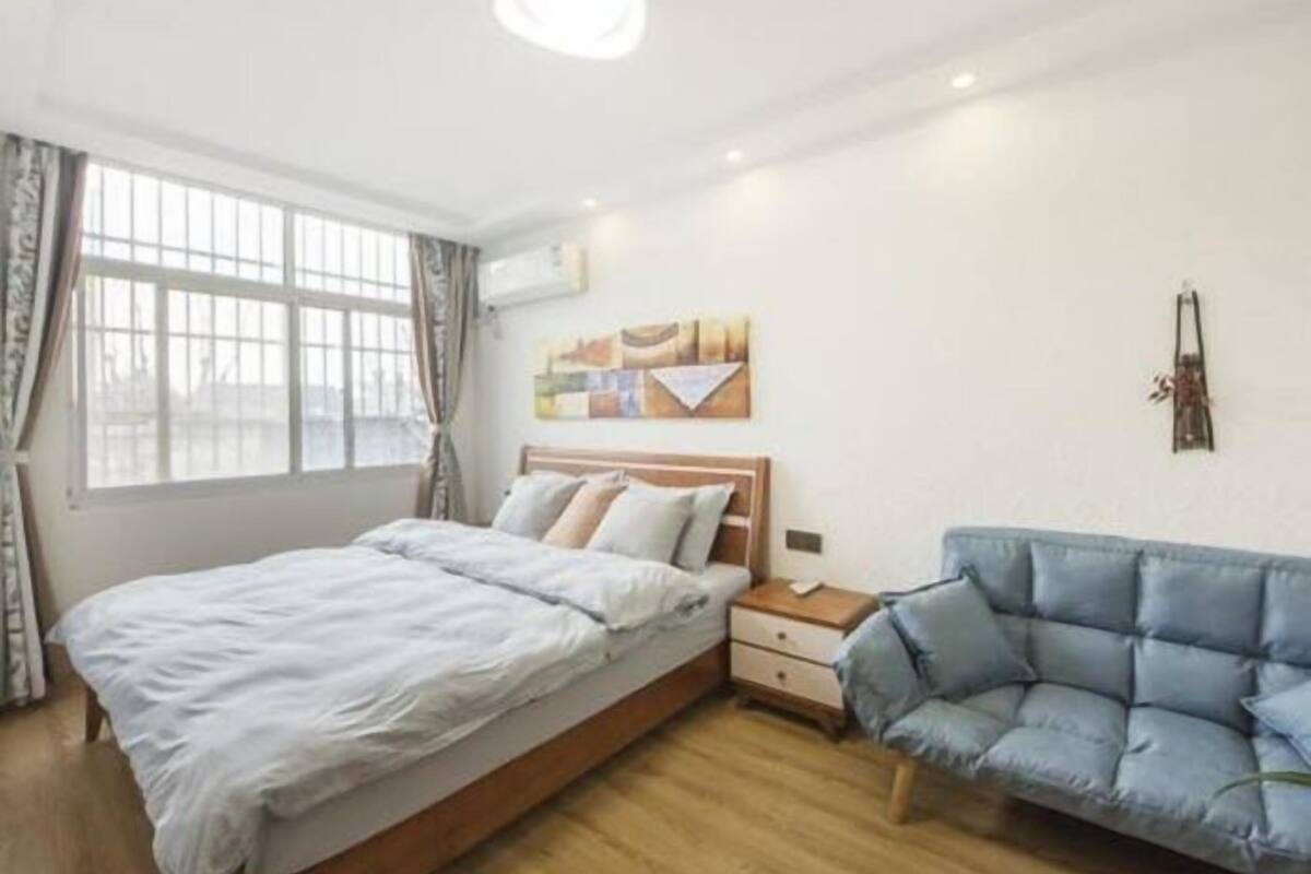 Quarto com cama, mesa de cabeceira, sofá azul, janela com cortinas e uma obra de arte na parede.