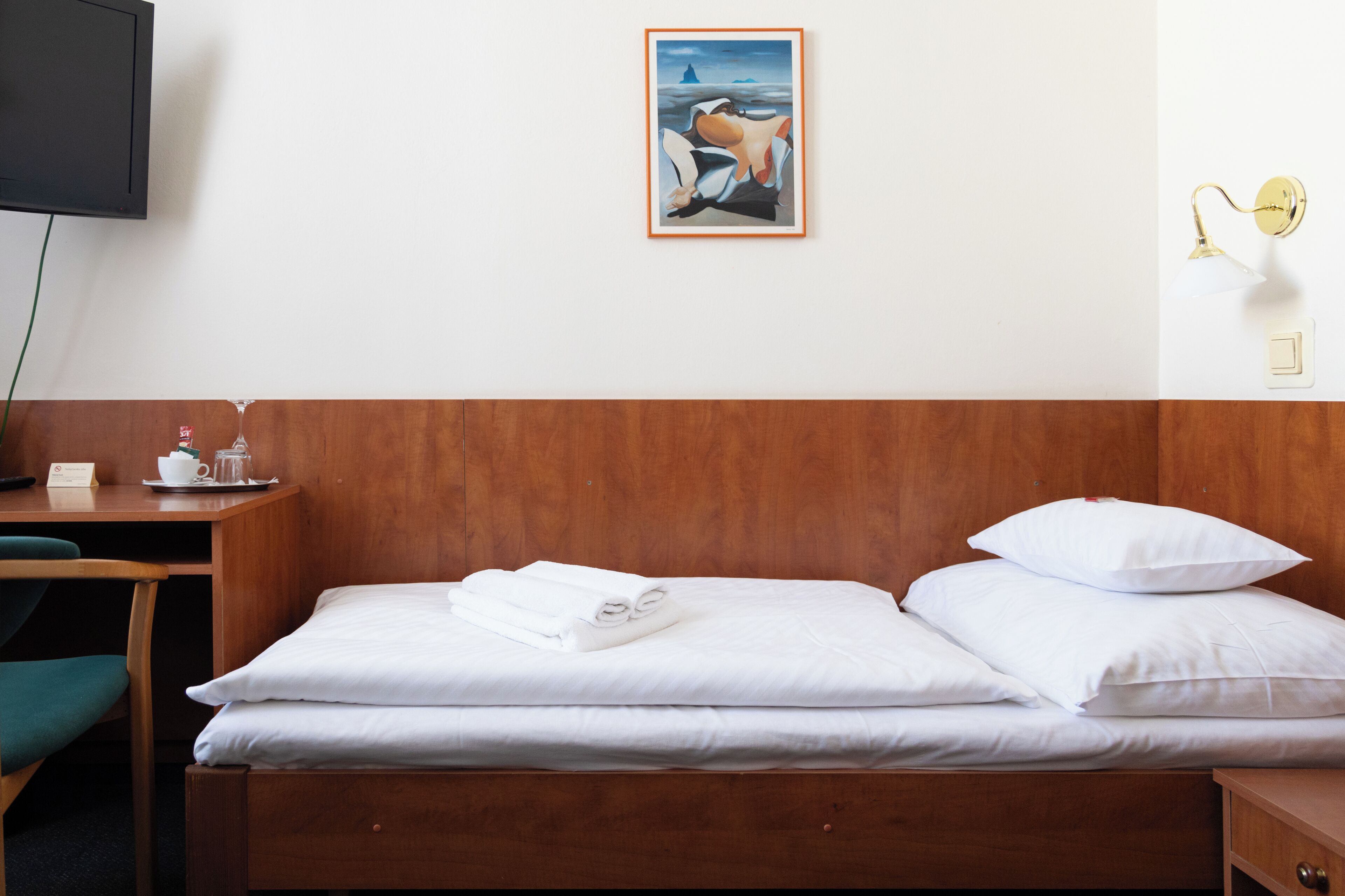Uma cama bem arrumada com lençóis brancos, uma cabeceira de madeira, uma obra de arte emoldurada na parede, uma escrivaninha com xícara e pires, e um abajur na mesa de cabeceira.