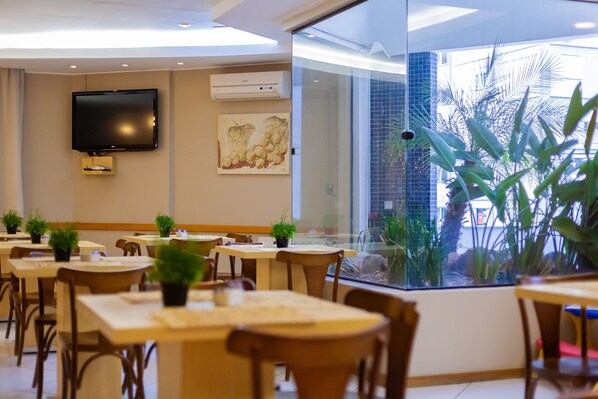 Um restaurante com várias mesas e cadeiras, uma televisão de tela plana na parede e uma janela grande com vista para palmeiras.