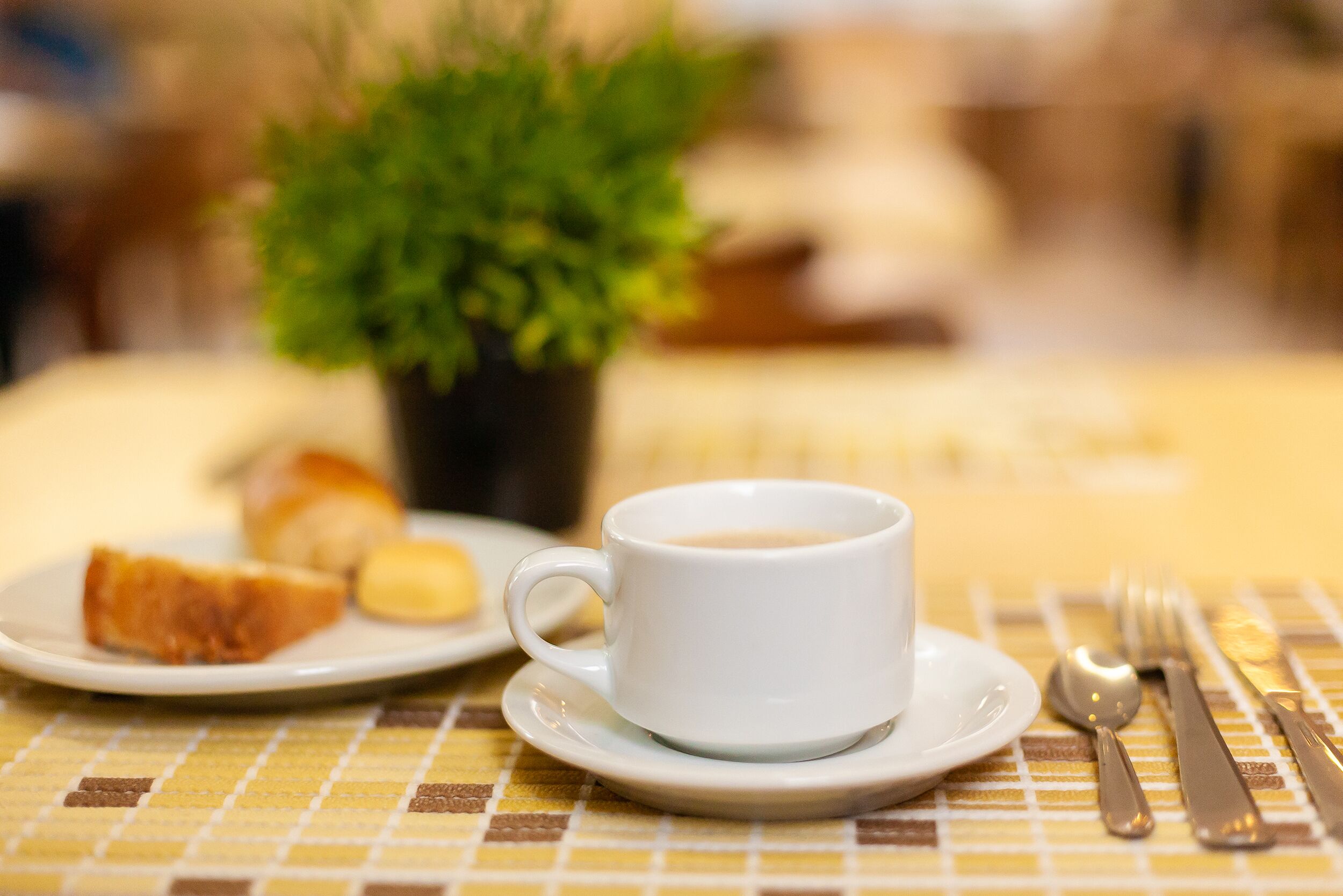 Uma mesa com uma xícara de café, um prato com croissant e manteiga, uma pequena planta e talheres.