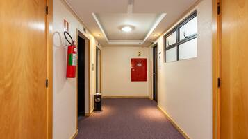 Corredor com portas de madeira, um extintor de incêndio, uma lixeira e uma placa de saída de emergência vermelha.