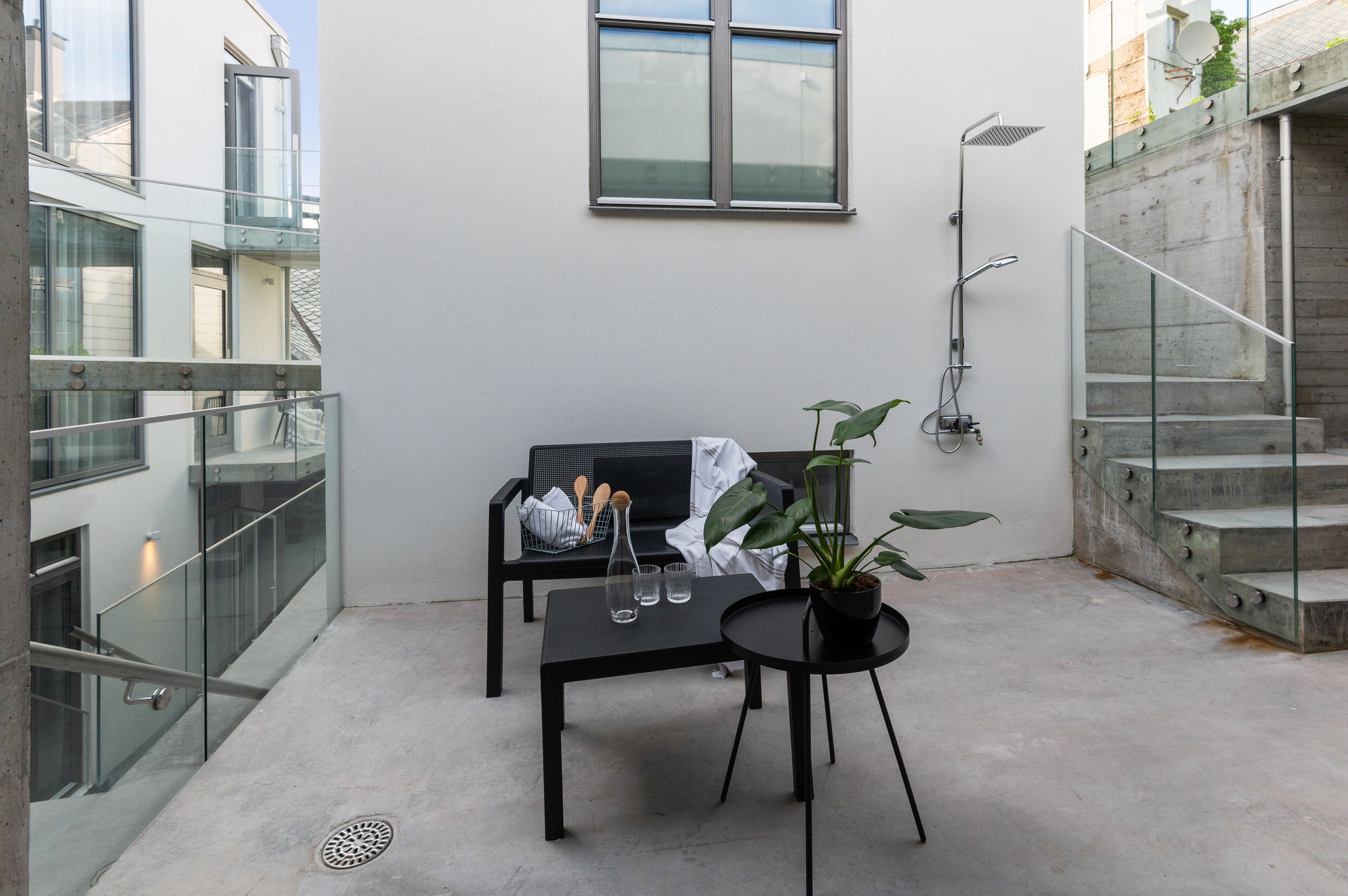 Uma varanda com guarda-corpo de vidro, uma mesa preta com planta em vaso e um banco.