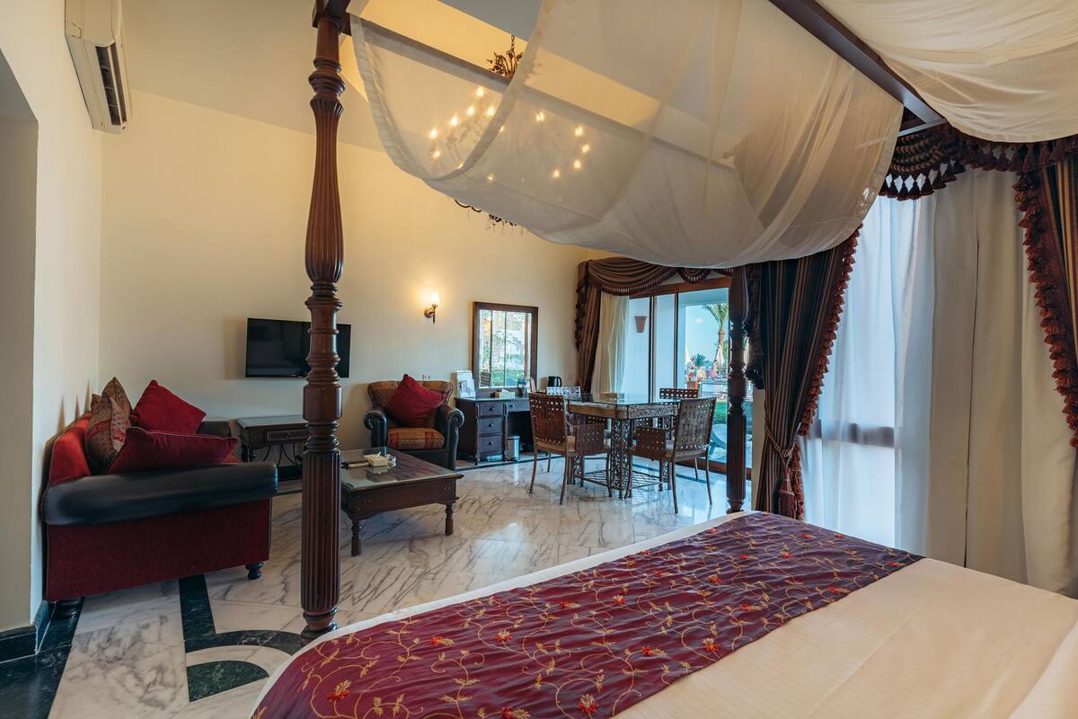 Quarto com cama de dossel, área de estar com sofá e poltrona, mesa de jantar e vista para o exterior.