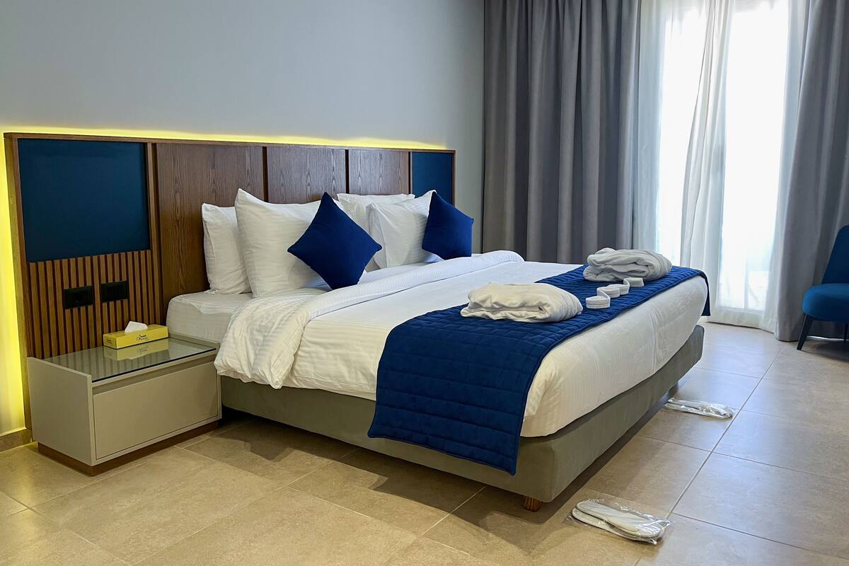 Quarto de hotel com uma cama, mesa de cabeceira, cadeira e janela com cortinas.