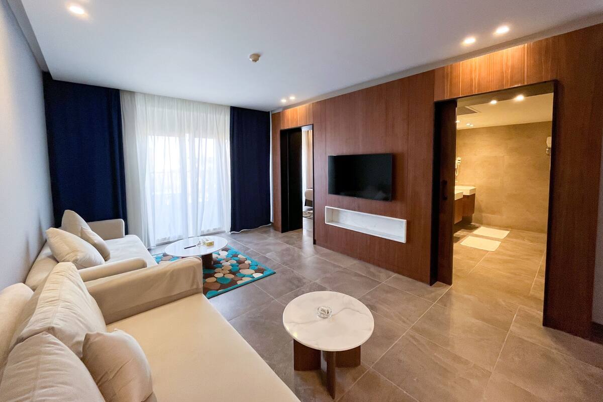 Uma sala de estar moderna com sofá, mesa de centro, TV na parede e um banheiro visível através de uma porta aberta.
