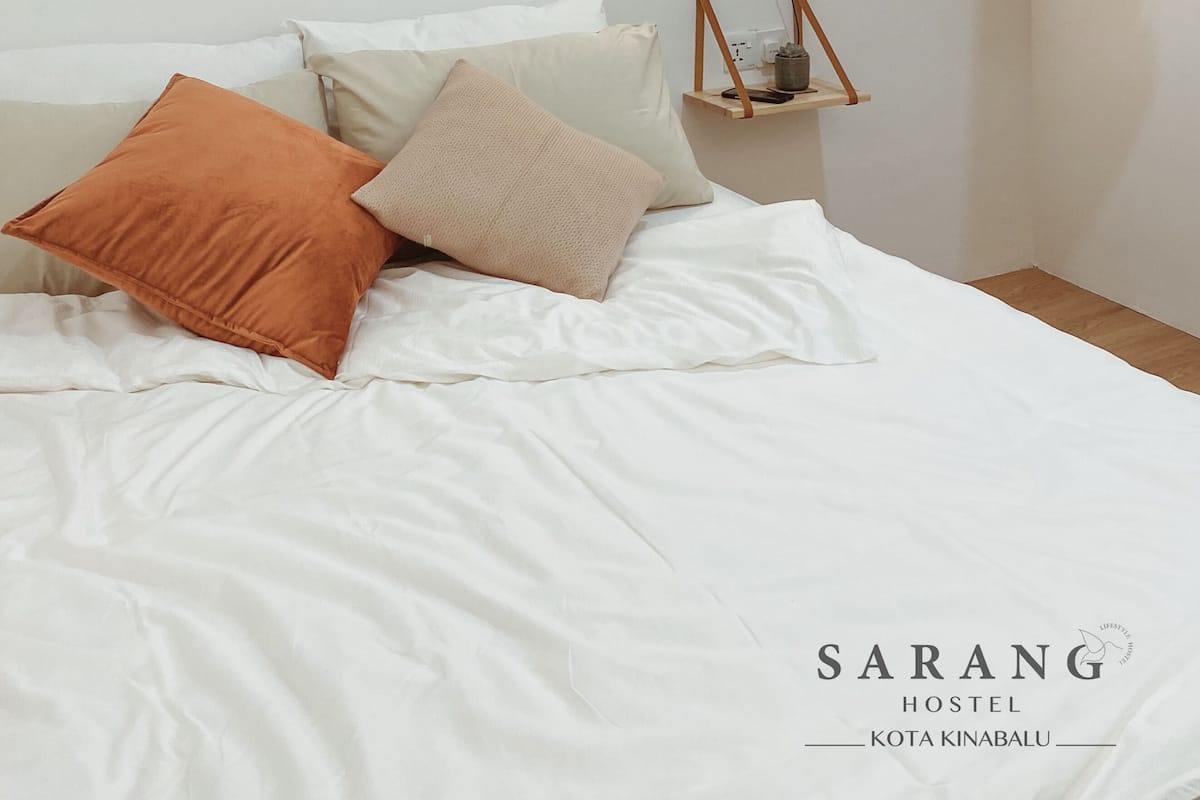 Uma cama bem arrumada com roupa de cama branca e duas almofadas, uma laranja e outra bege. Uma luminária de parede com base de madeira e um pendente de macramé são visíveis. O quarto tem um design minimalista, com um esquema de cores limpo e branco.
