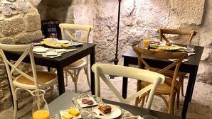 Ambiente de café da manhã com paredes de pedra, cadeiras de madeira e uma mesa posta com pratos de comida, copos e um livro intitulado "LA MIA SICILIA".