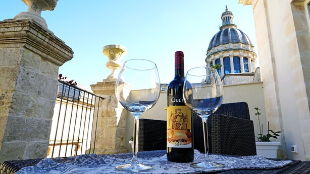 Uma garrafa de vinho e duas taças sobre uma mesa com toalha rendada, com uma arquitetura clássica e céu claro ao fundo.