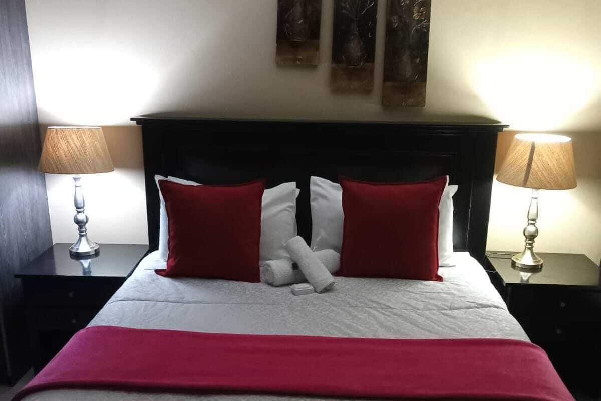 Cama bem arrumada com esquema de cores vermelho e branco, ladeada por duas mesas de cabeceira com abajures, e três obras de arte penduradas na parede acima da cama.