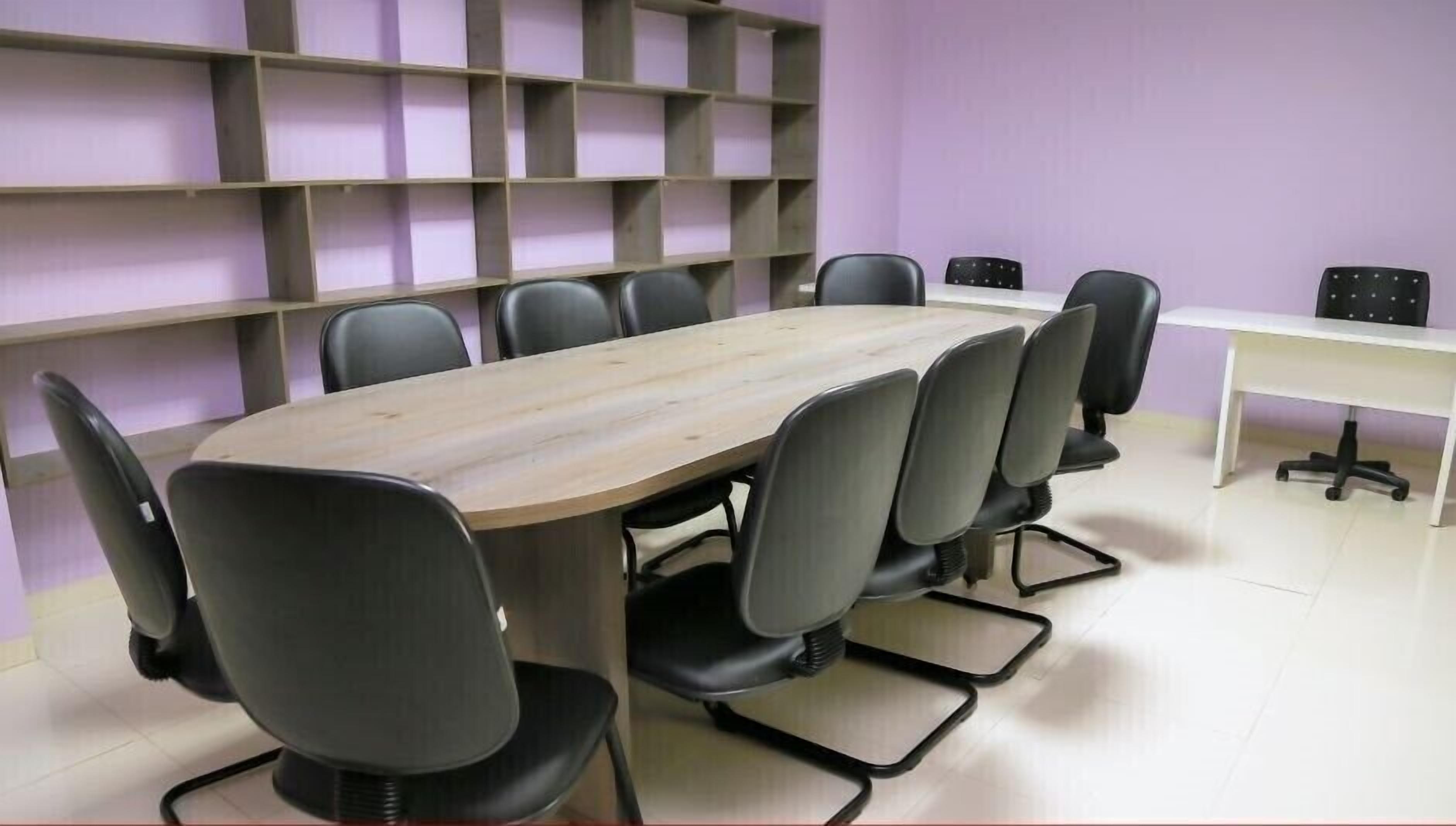 Uma sala de conferências com uma mesa comprida e várias cadeiras, uma escrivaninha branca e estantes na parede.