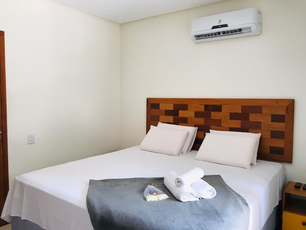 Quarto de hotel com uma cama, cabeceira de madeira, ar condicionado branco e uma pequena mesa com um controle remoto.