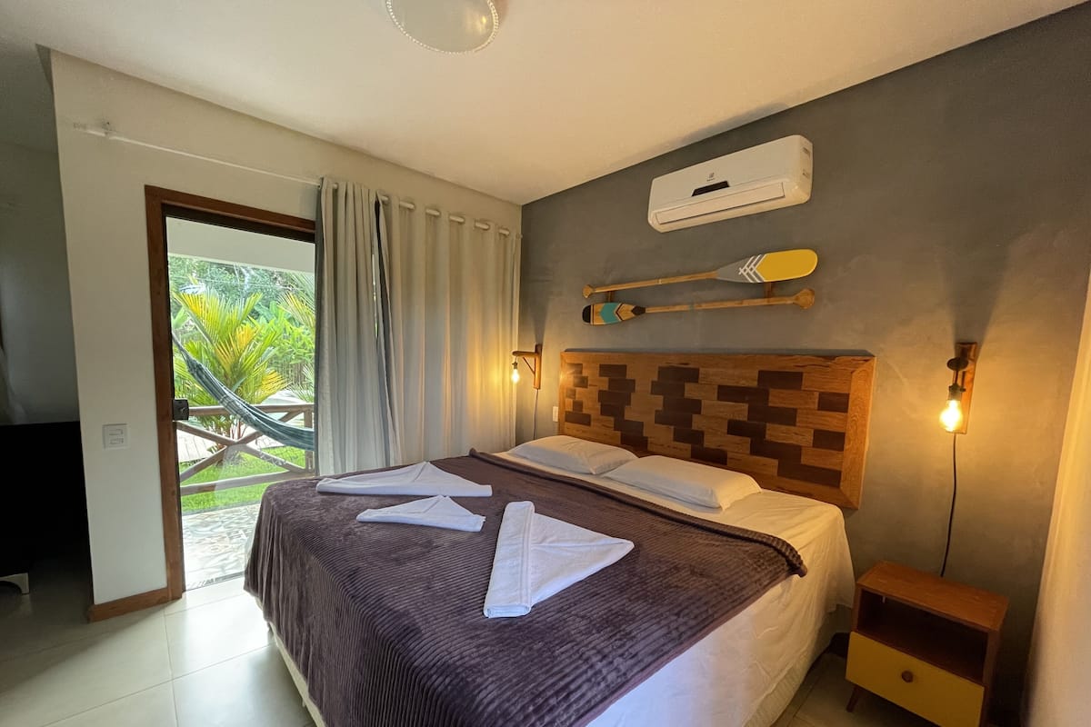 Quarto de hotel com cama, cabeceira de madeira, mesa de cabeceira, ar condicionado fixado na parede e vista para o exterior.