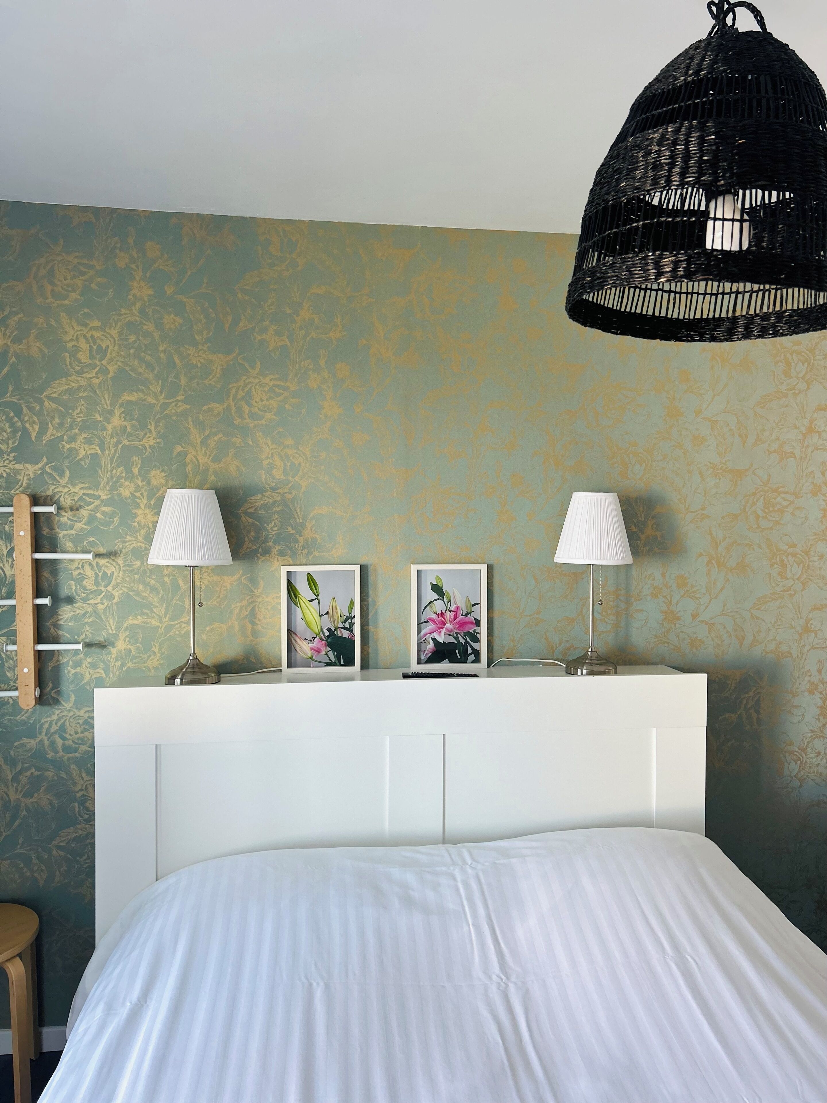 Quarto com cama branca, duas luminárias de parede e duas imagens florais emolduradas sobre um console branco.